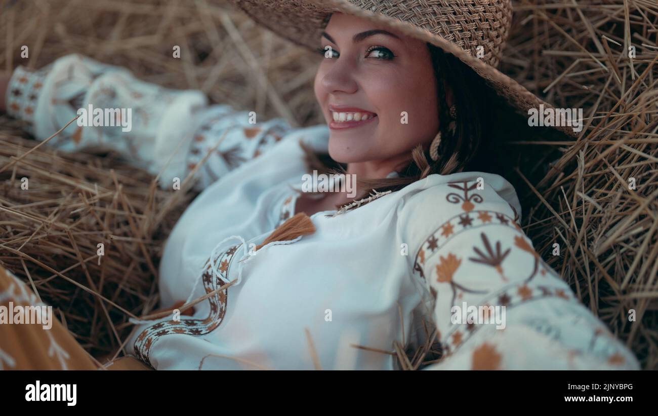 Pretty woman in straw hat and embroidered blouse smiling lying on hay ...