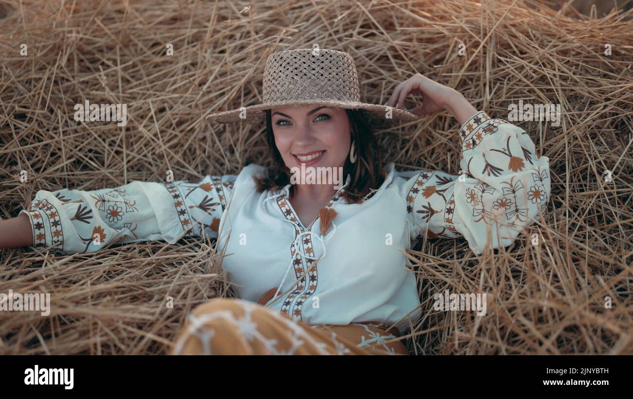 Pretty woman in straw hat and embroidered blouse smiling lying on hay ...