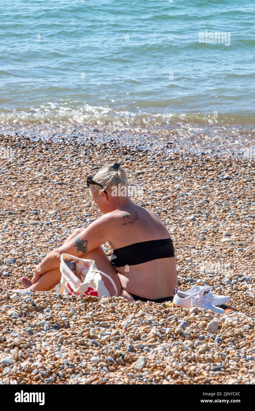 young blond woman sitting on a pebble beach in the hot summer sunshine during a heatwave and ...