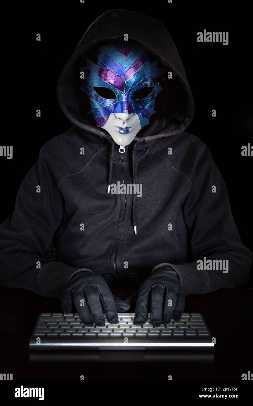 A hooded person with a mask, typing on a computer keyboard. Low key ...