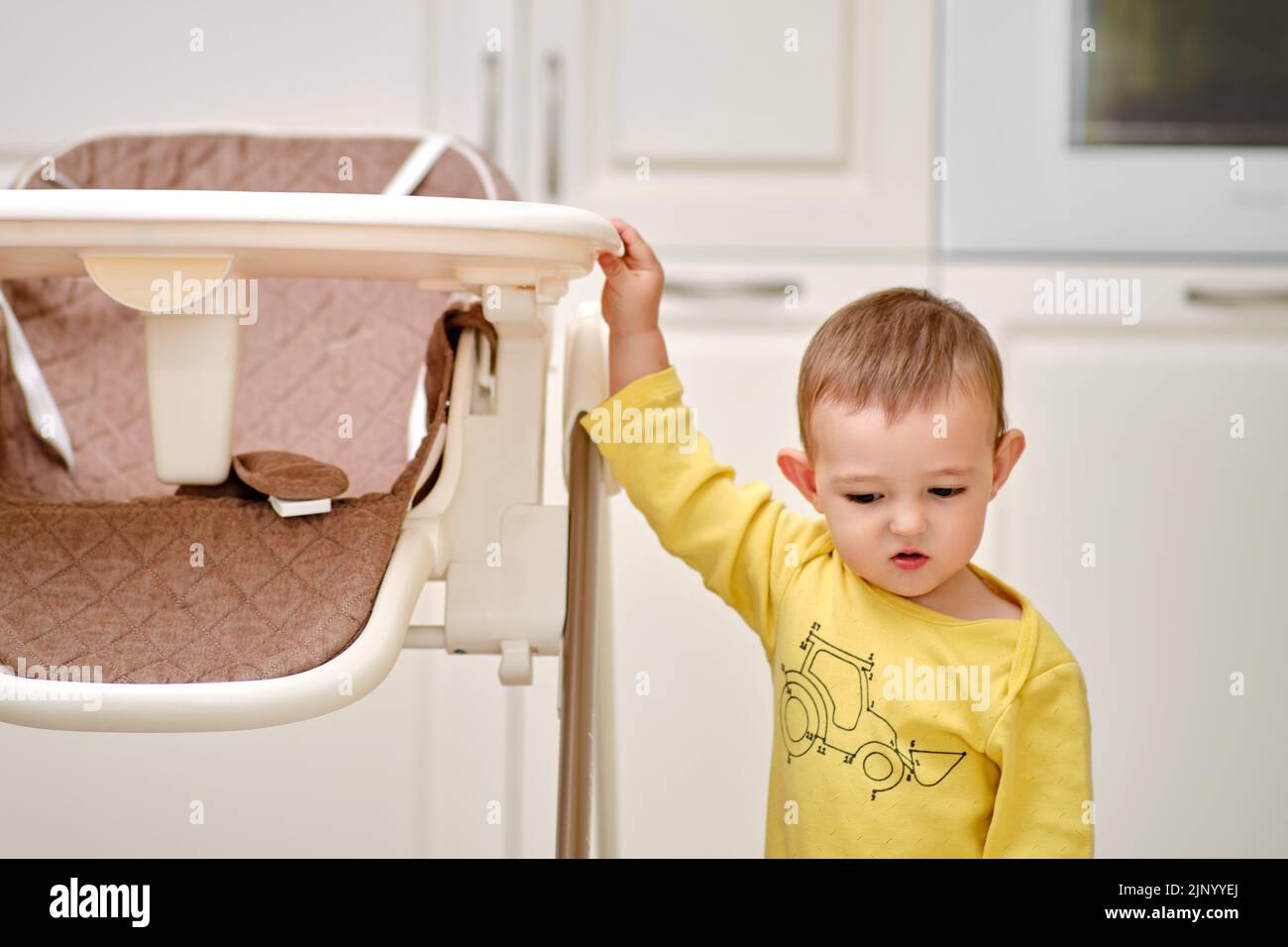 Sad toddler baby boy stands holding onto a children's high chair in the ...