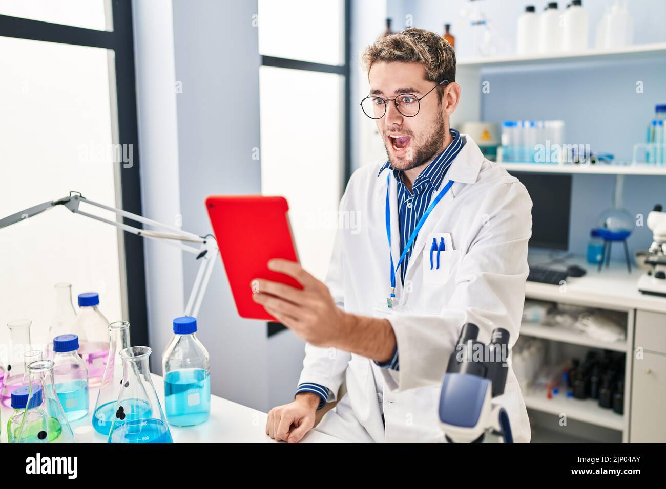 Hispanic man with beard working at scientist laboratory doing video call scared and amazed with ...