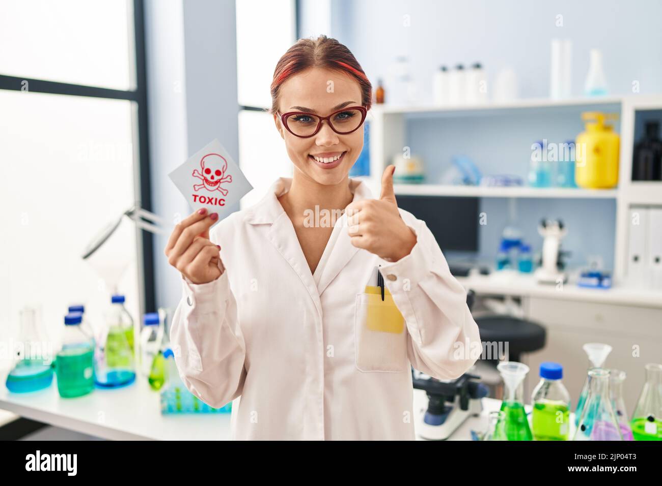 Young caucasian woman working at scientist laboratory holding toxic label smiling happy and ...