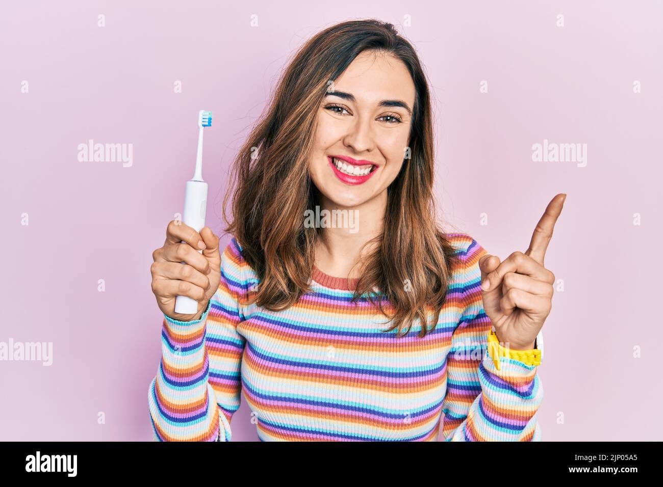 Young hispanic girl holding electric toothbrush surprised with an idea or question pointing ...