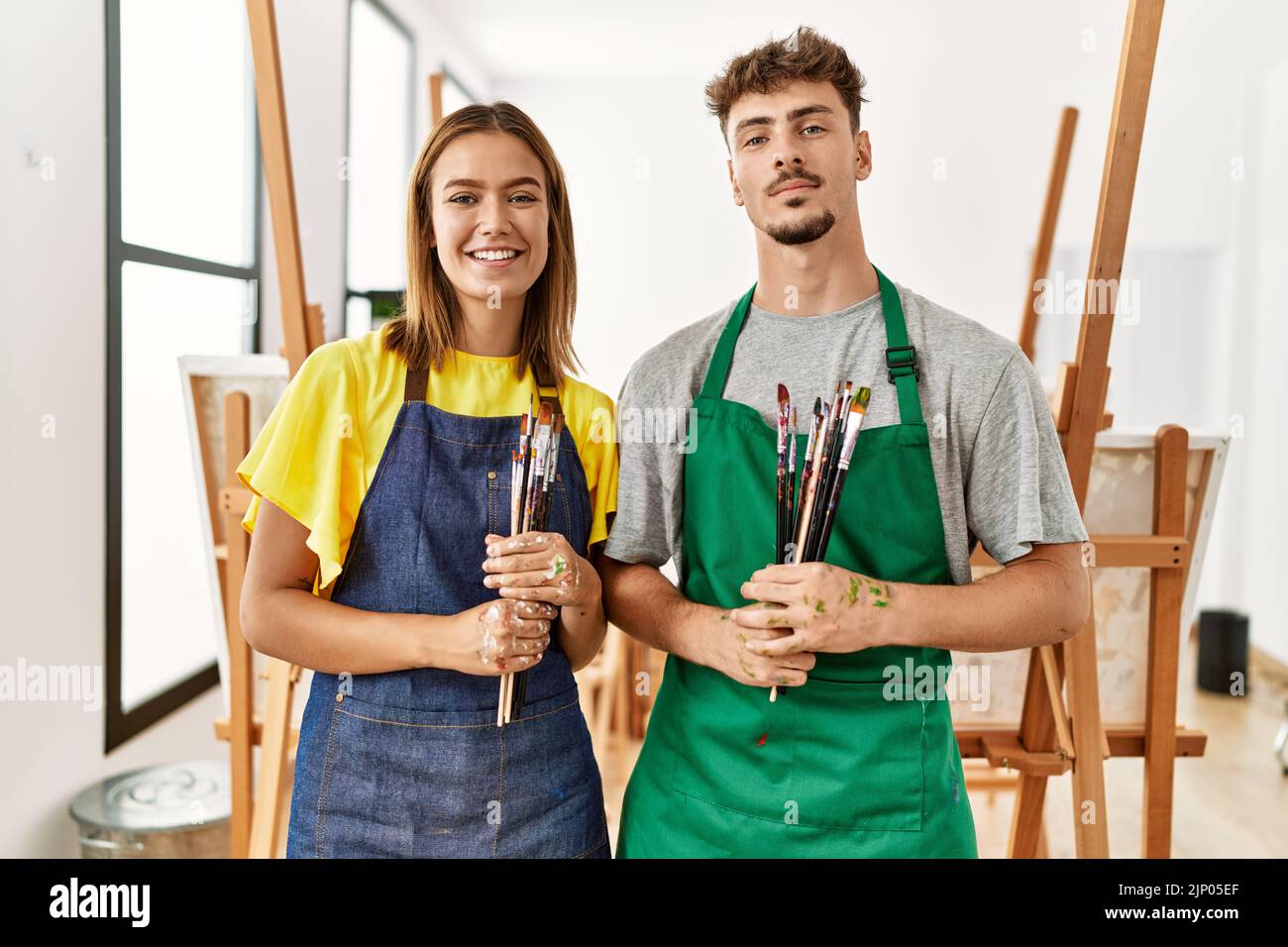 Young hispanic artist couple smiling happy holding paintbrushes at art ...