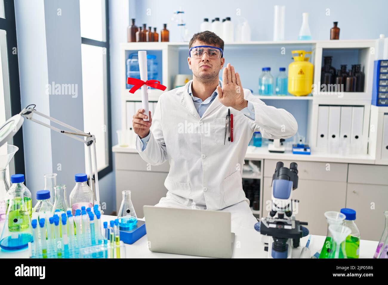 Young hispanic man working at scientist laboratory holding diploma with ...