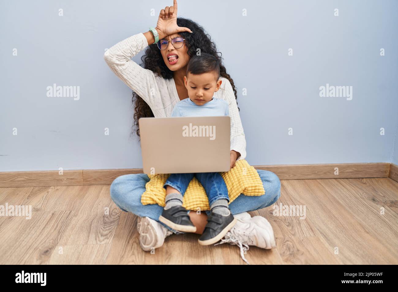 Young hispanic mother and kid using computer laptop sitting on the floor making fun of people ...
