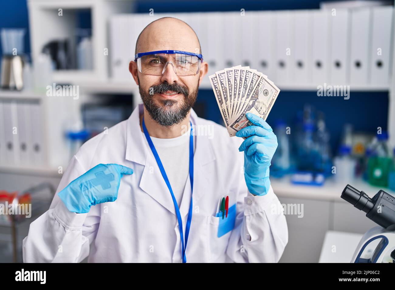 Young hispanic man working at scientist laboratory holding money pointing finger to one self ...