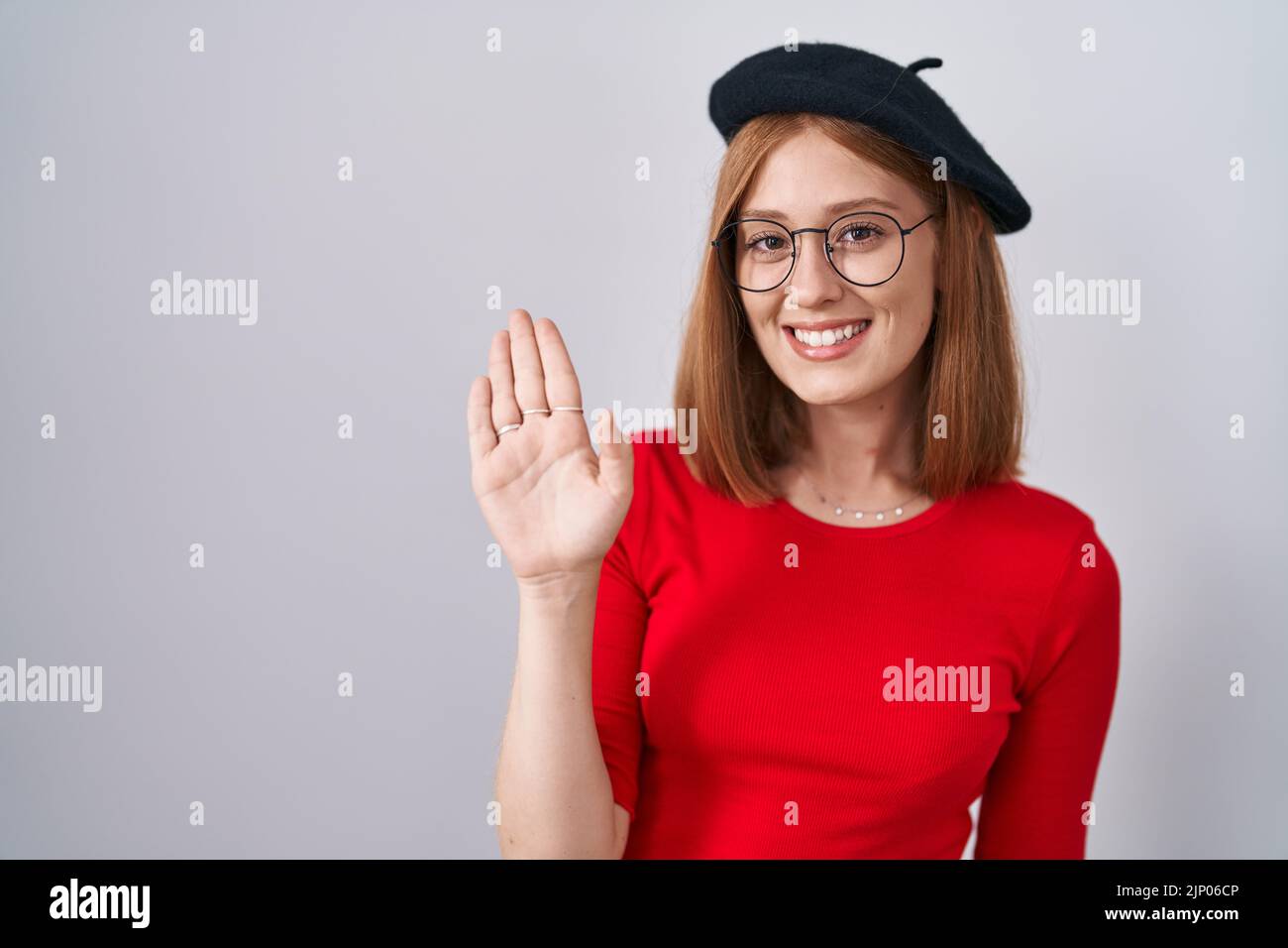 Young redhead woman standing wearing glasses and beret waiving saying ...