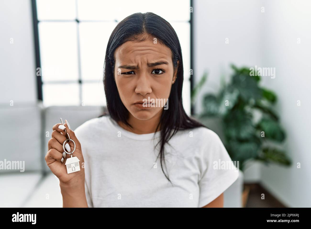Young hispanic woman holding keys of new home skeptic and nervous, frowning upset because of ...