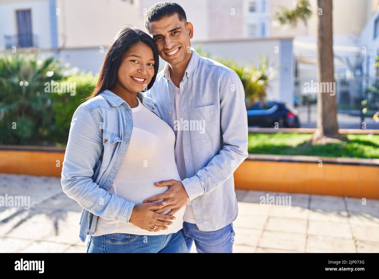 Young latin couple expecting baby hugging each other standing at park ...