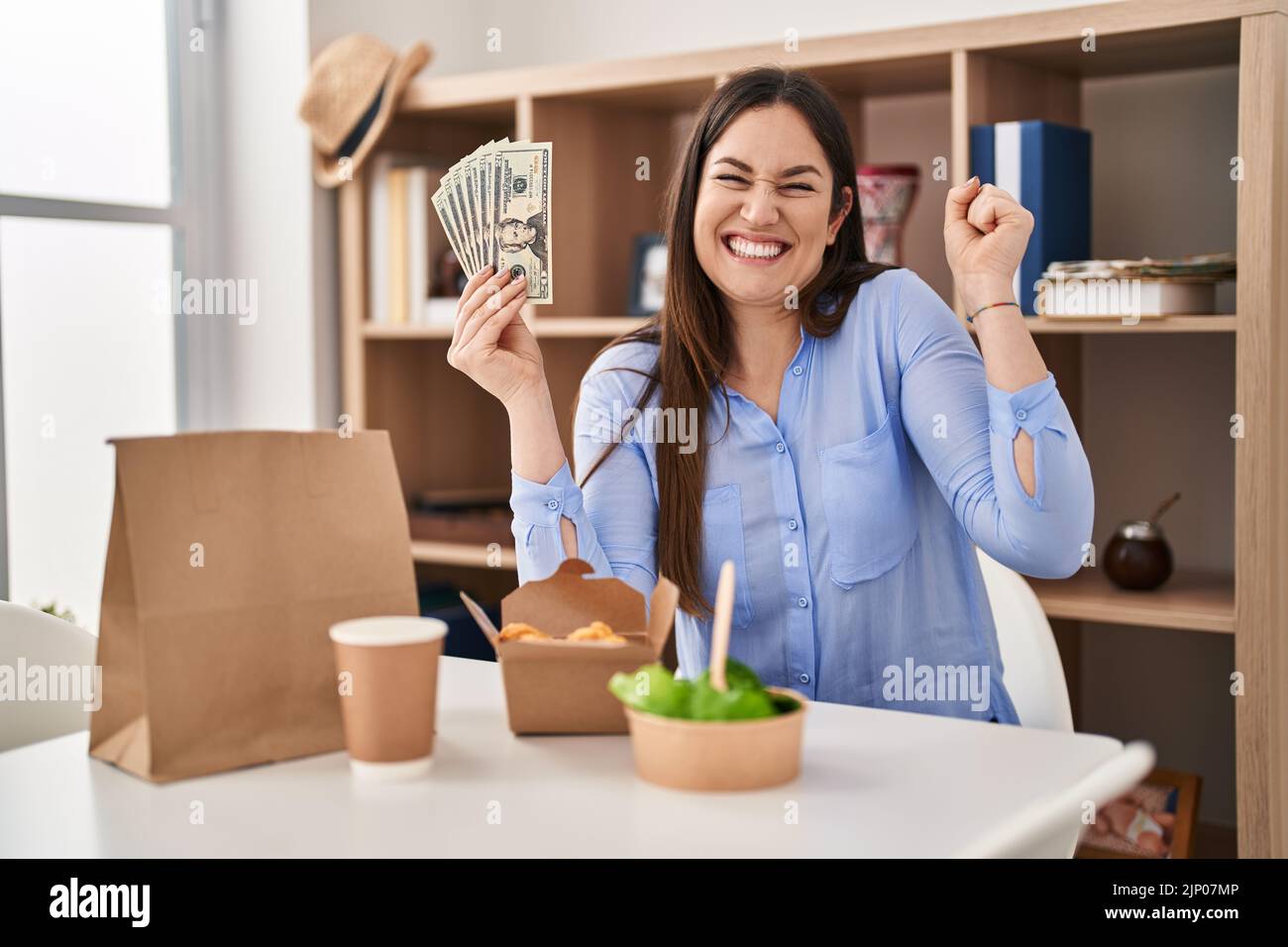 Young Brunette Woman Eating Take Away Food At Home Holding Money