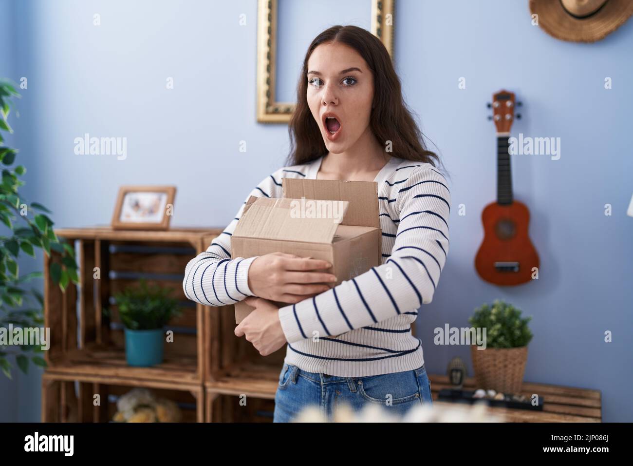 Young hispanic girl opening cardboard box afraid and shocked with ...