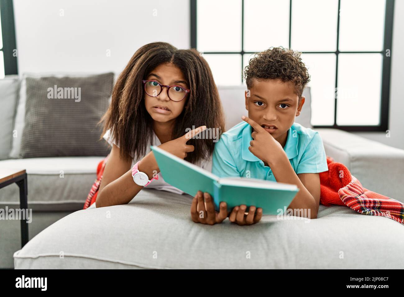 Two siblings lying on the sofa reading a book pointing aside worried ...