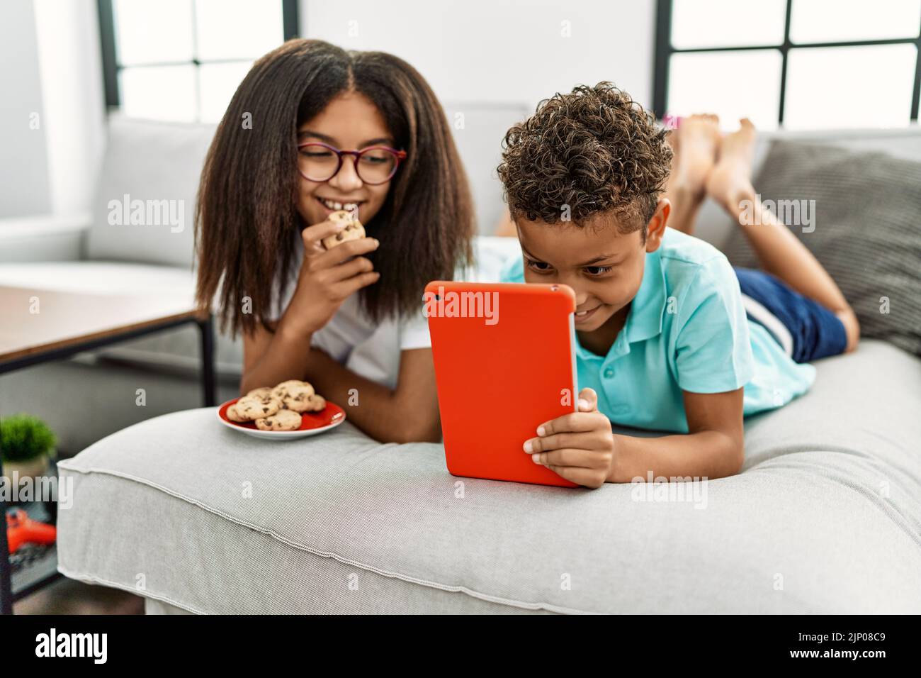 Brother and sister eating cookies using touchpad lying on sofa at home ...