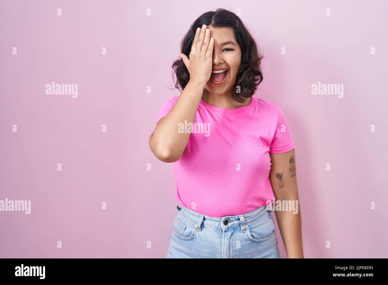 Young hispanic woman standing over pink background covering one eye ...
