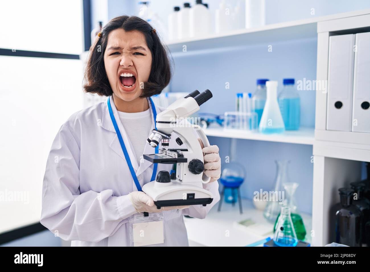 Young hispanic woman working at scientist laboratory holding microscope ...