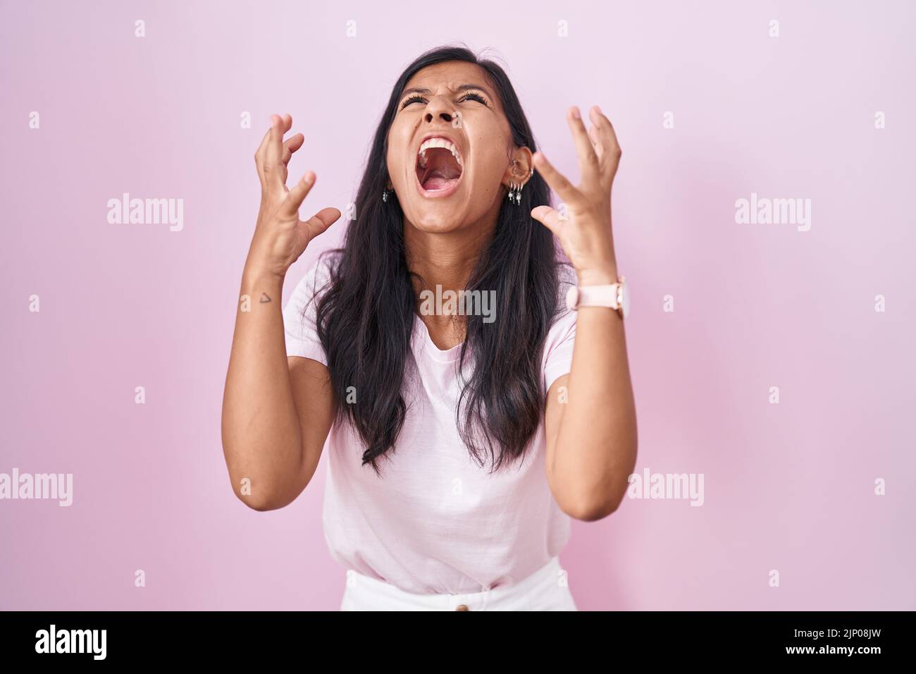 Young hispanic woman standing over pink background crazy and mad ...