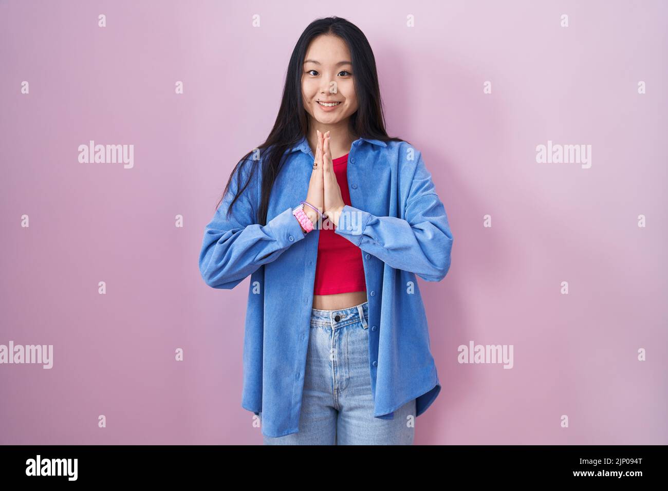 Young asian woman standing over pink background praying with hands ...
