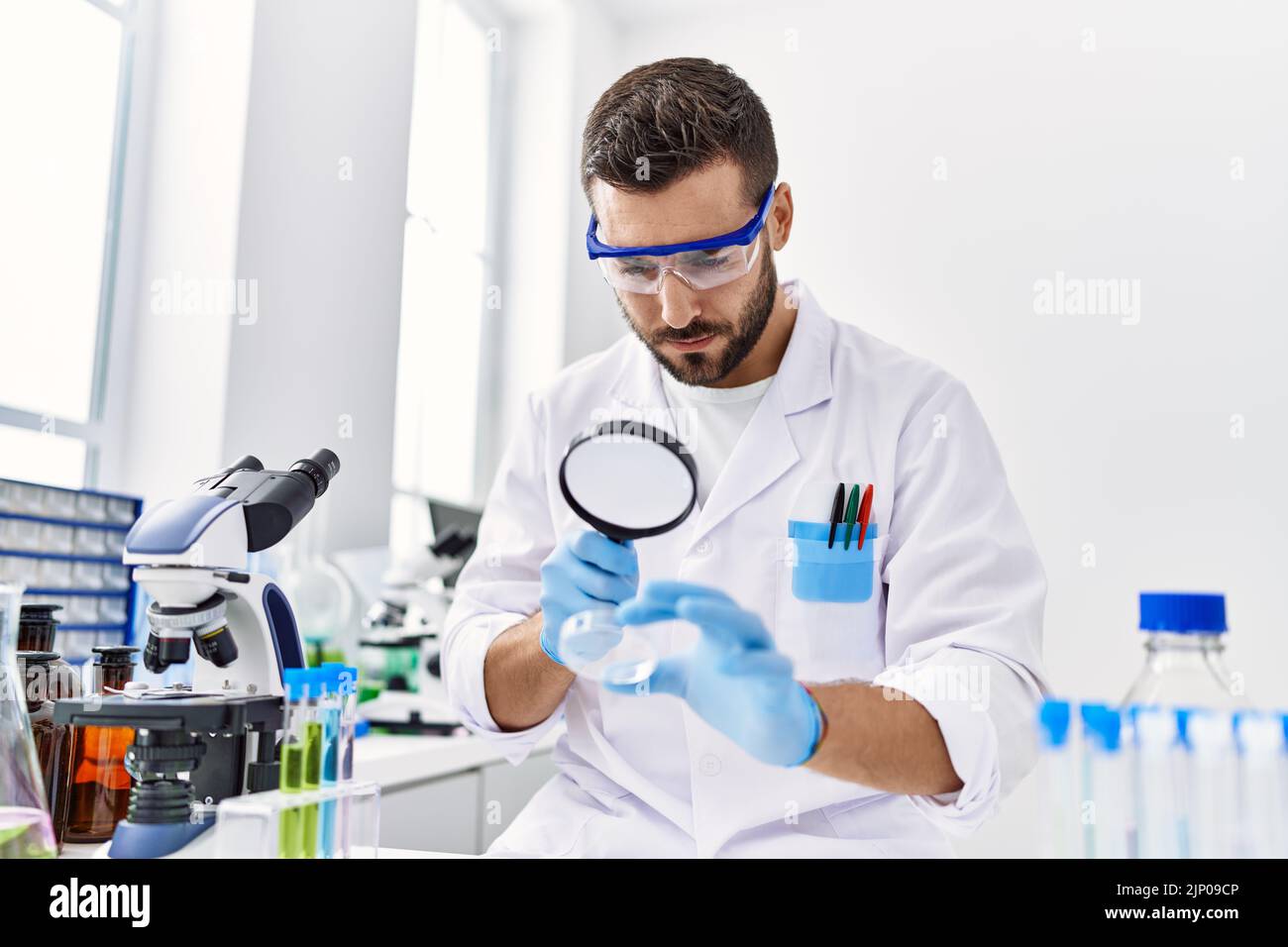 Young hispanic man wearing scientist uniform using loupe at laboratory ...