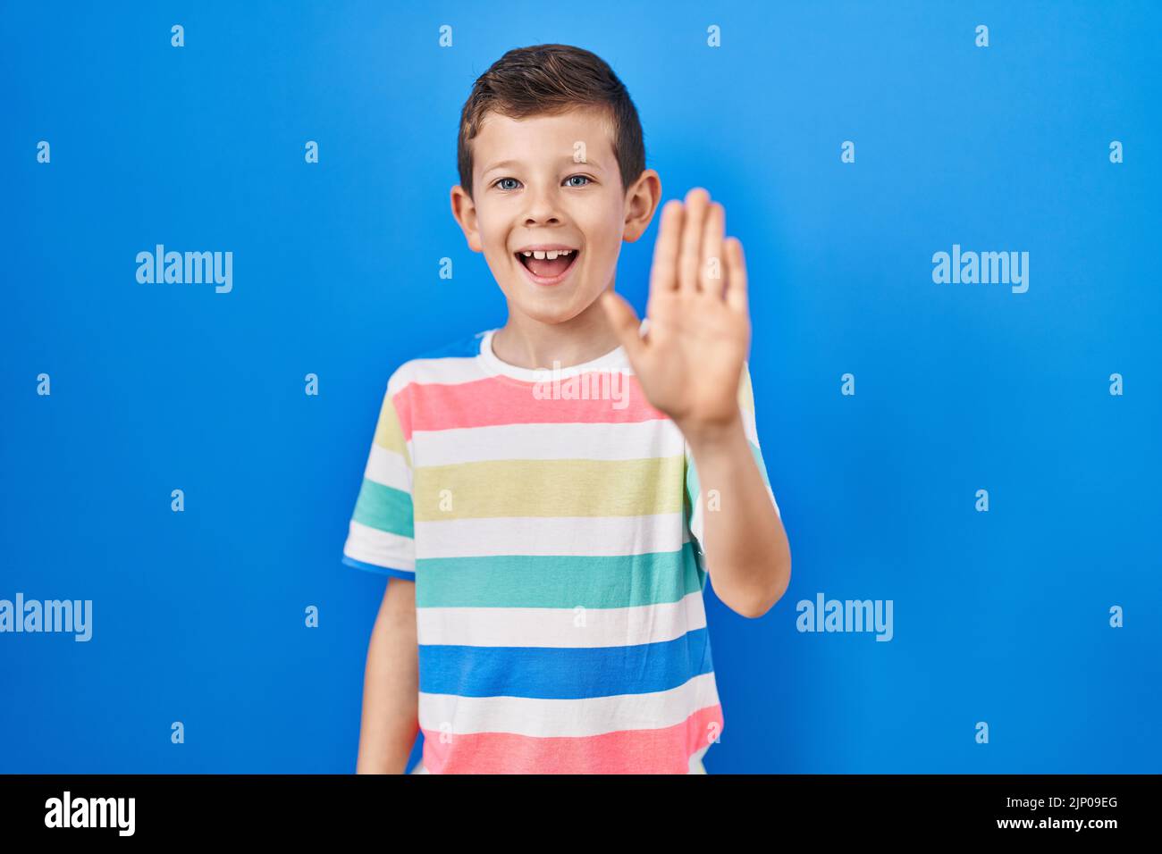 Young caucasian kid standing over blue background waiving saying hello ...
