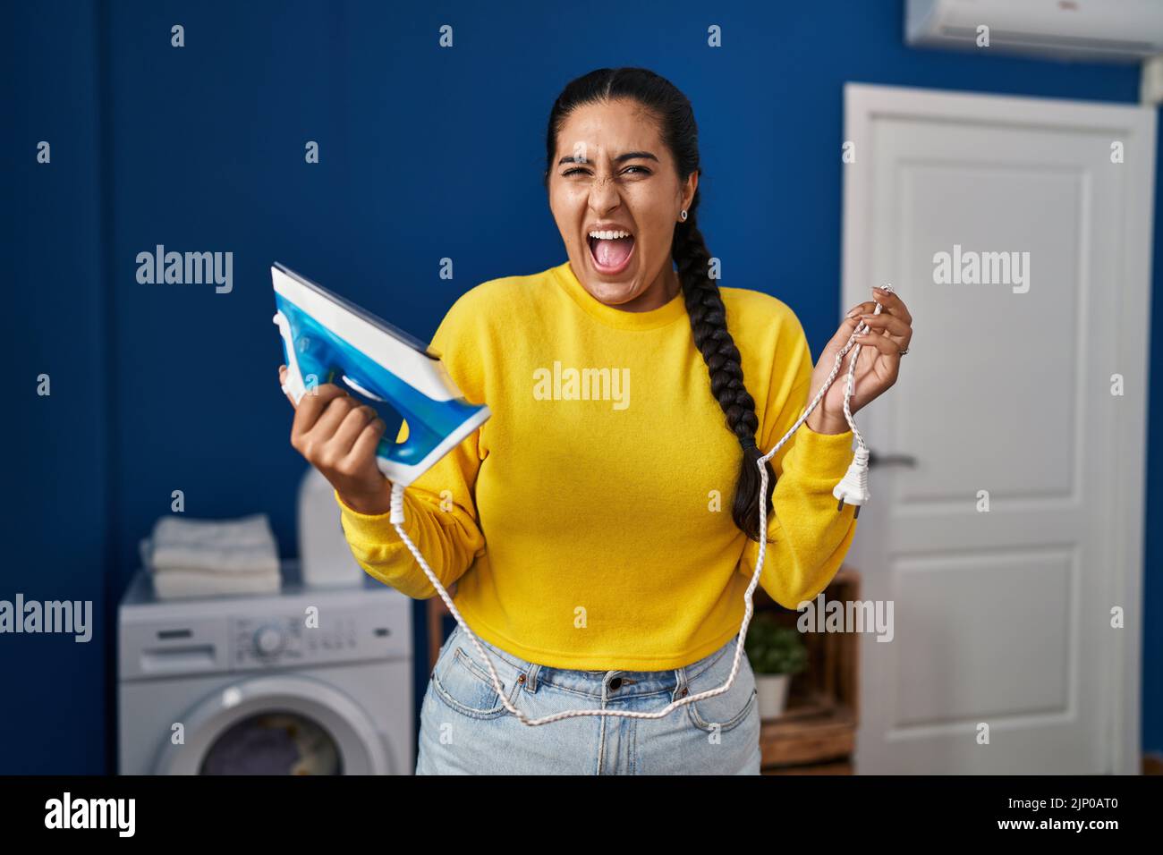 Young hispanic woman holding iron at laundry room angry and mad ...