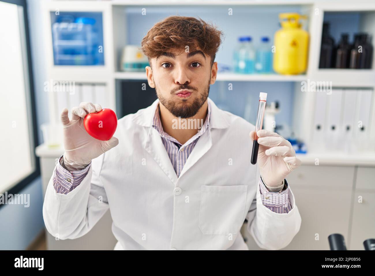 Arab man with beard working at scientist laboratory holding blood samples puffing cheeks with ...