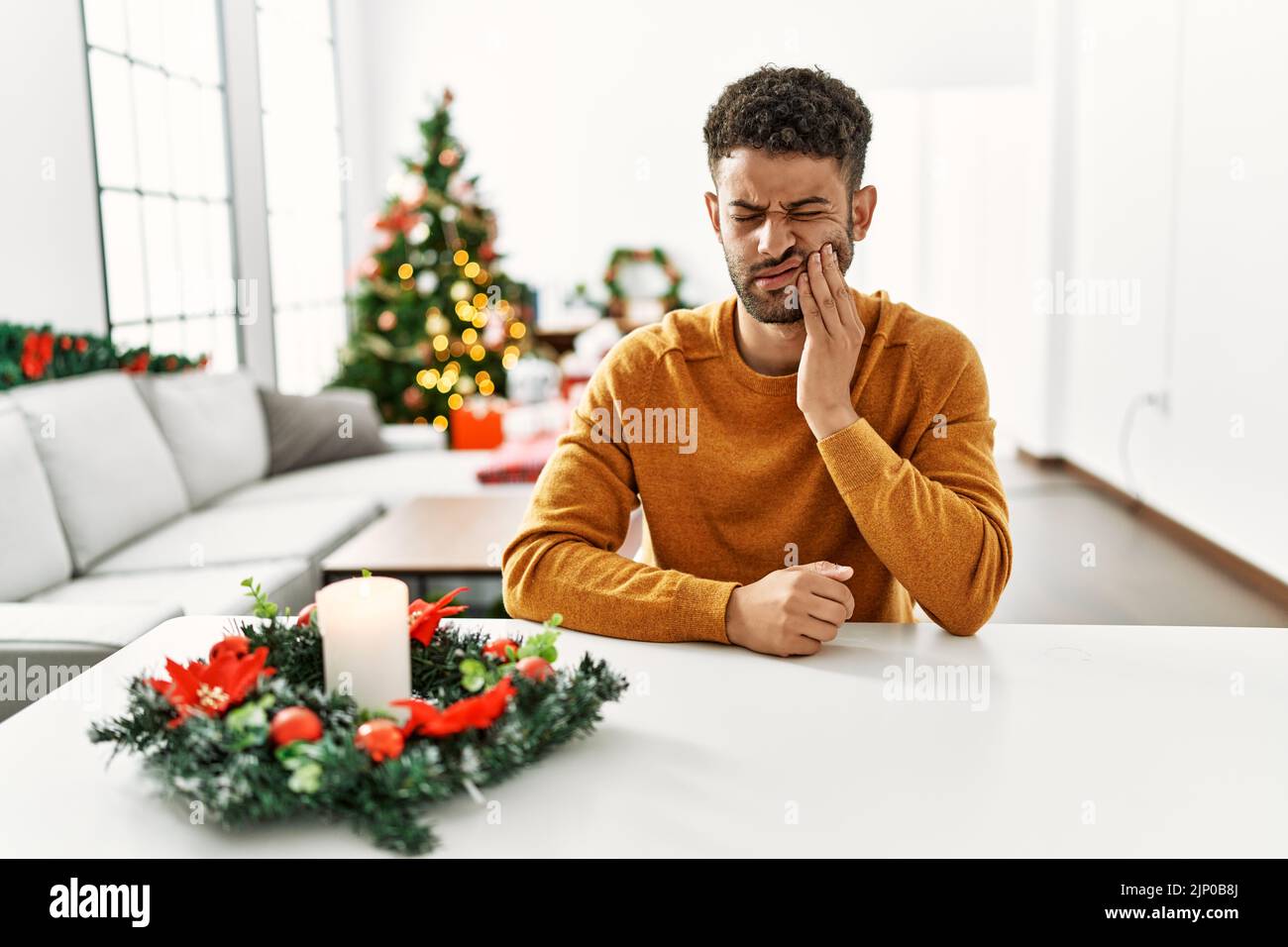 Arab young man sitting on the table by christmas tree touching mouth ...