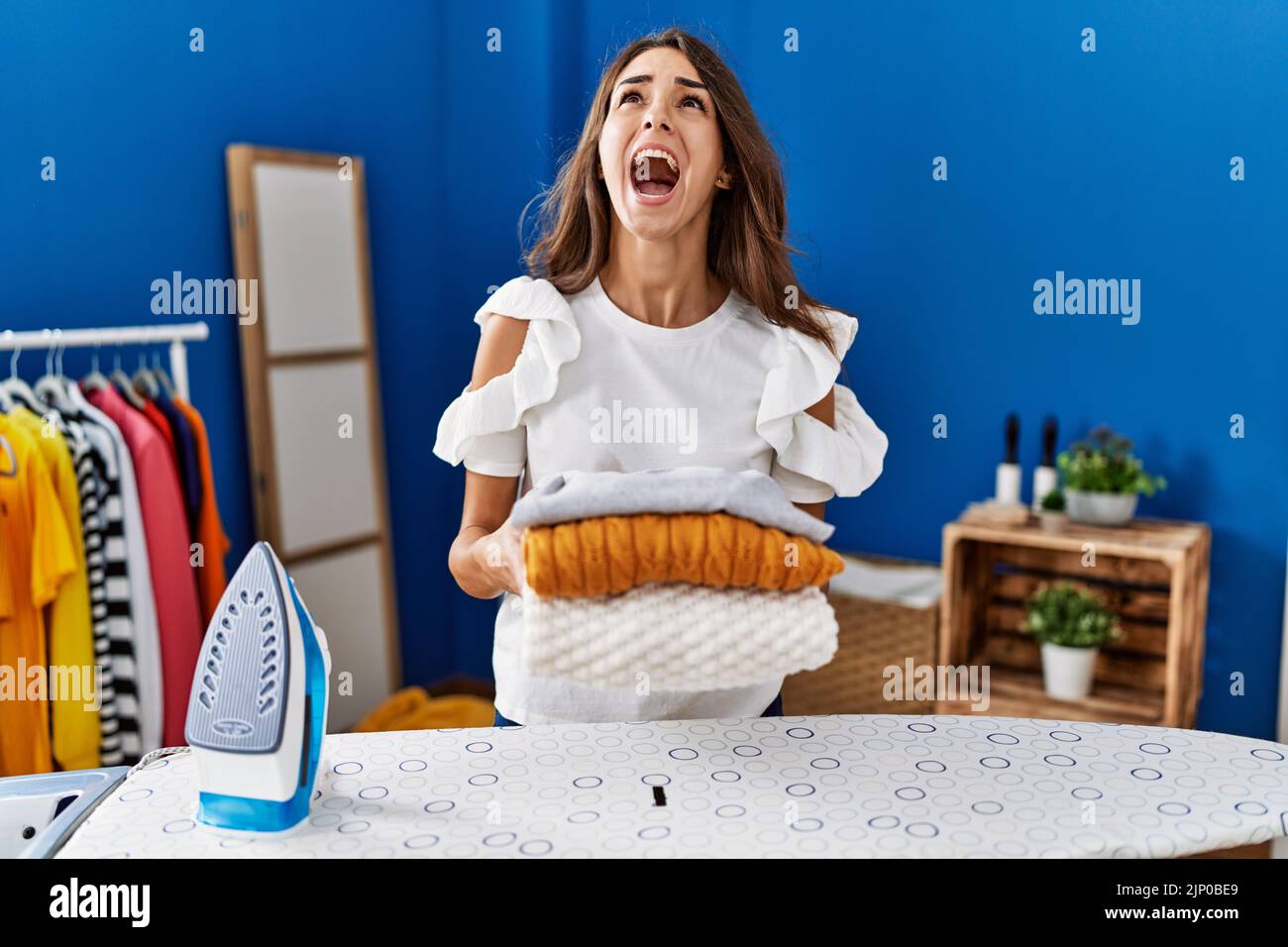 Young hispanic woman ironing clothes at laundry room holding folded ...