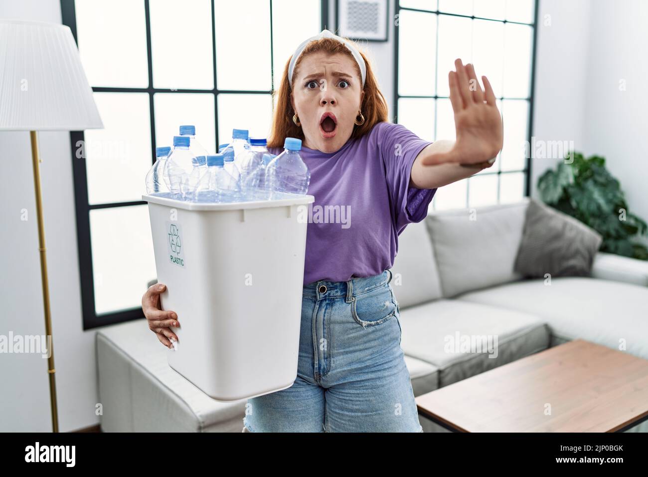 Young redhead woman holding recycling wastebasket with plastic bottles ...