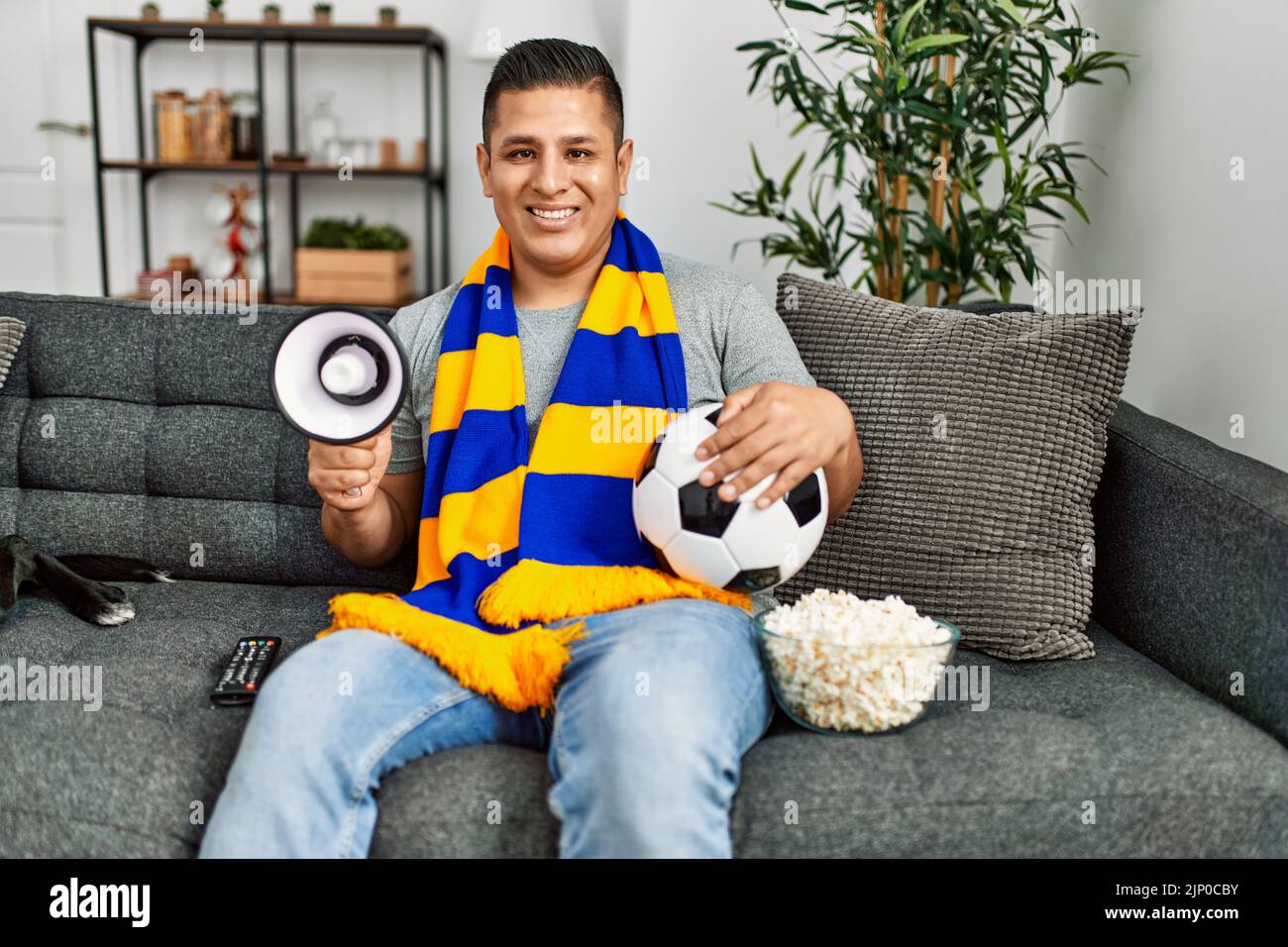 Young hispanic hooligan man supporting soccer team sitting on the sofa ...