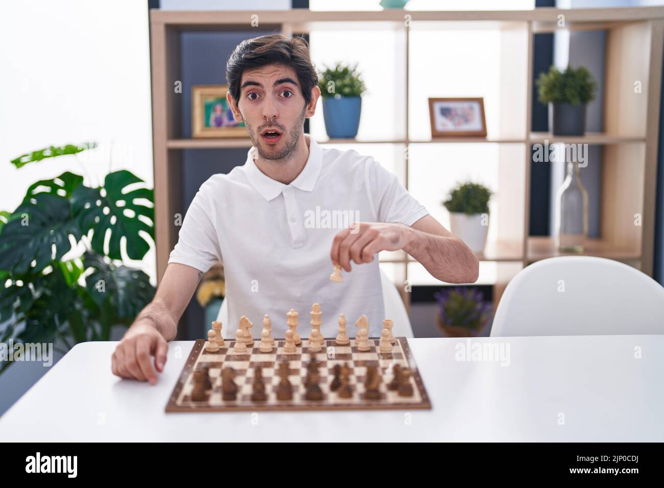 Young hispanic man playing chess sitting on the table scared and amazed ...