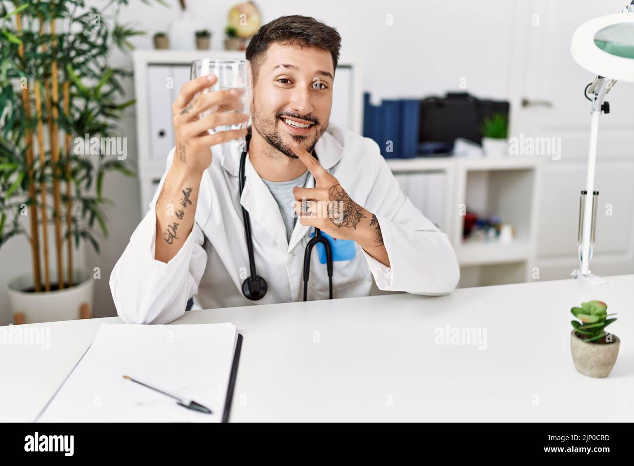Young doctor holding glass of water smiling happy pointing with hand ...