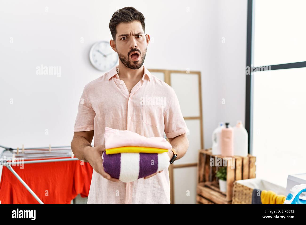 Young man with beard holding folded laundry after ironing in shock face ...