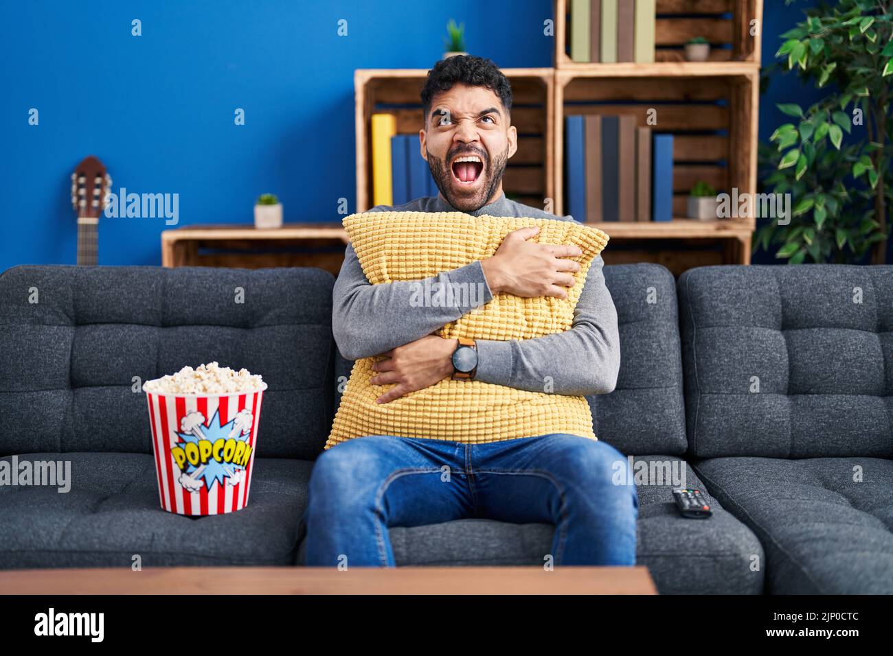 Hispanic man with beard eating popcorn watching a movie at home angry ...