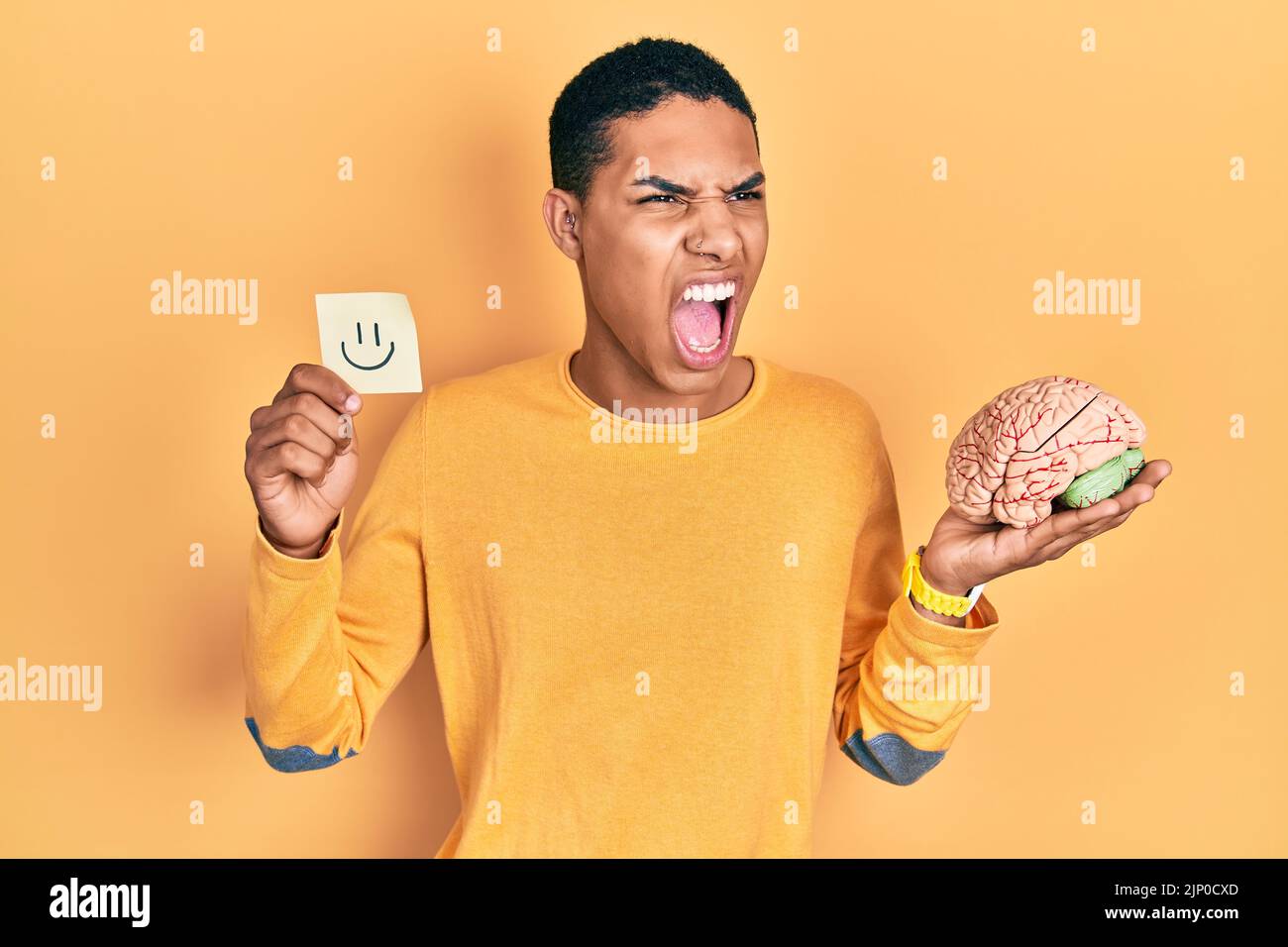 Young african american guy holding smile reminder and brain angry and ...