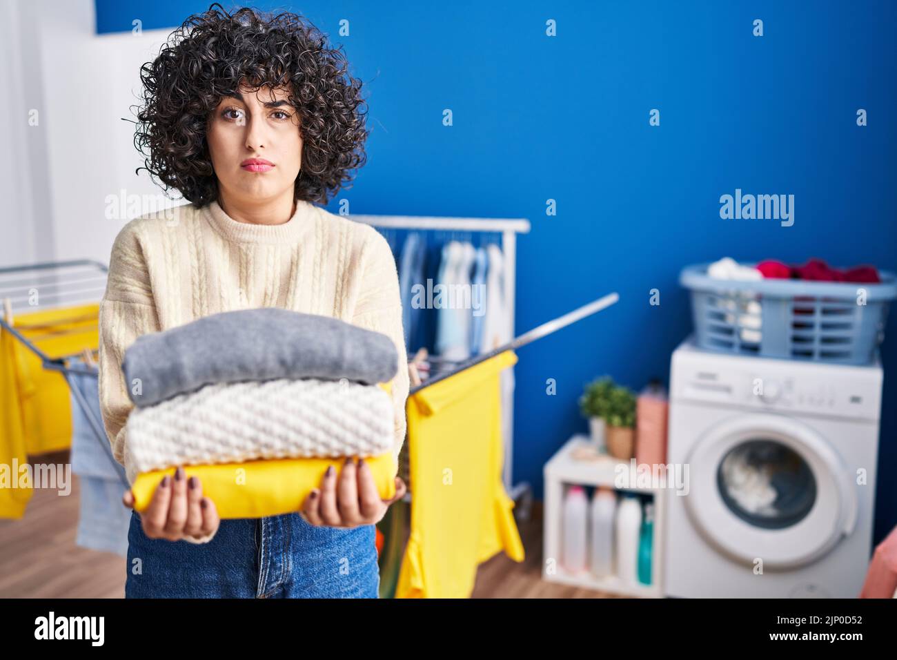 Young brunette woman with curly hair holding clean laundry skeptic and nervous, frowning upset ...