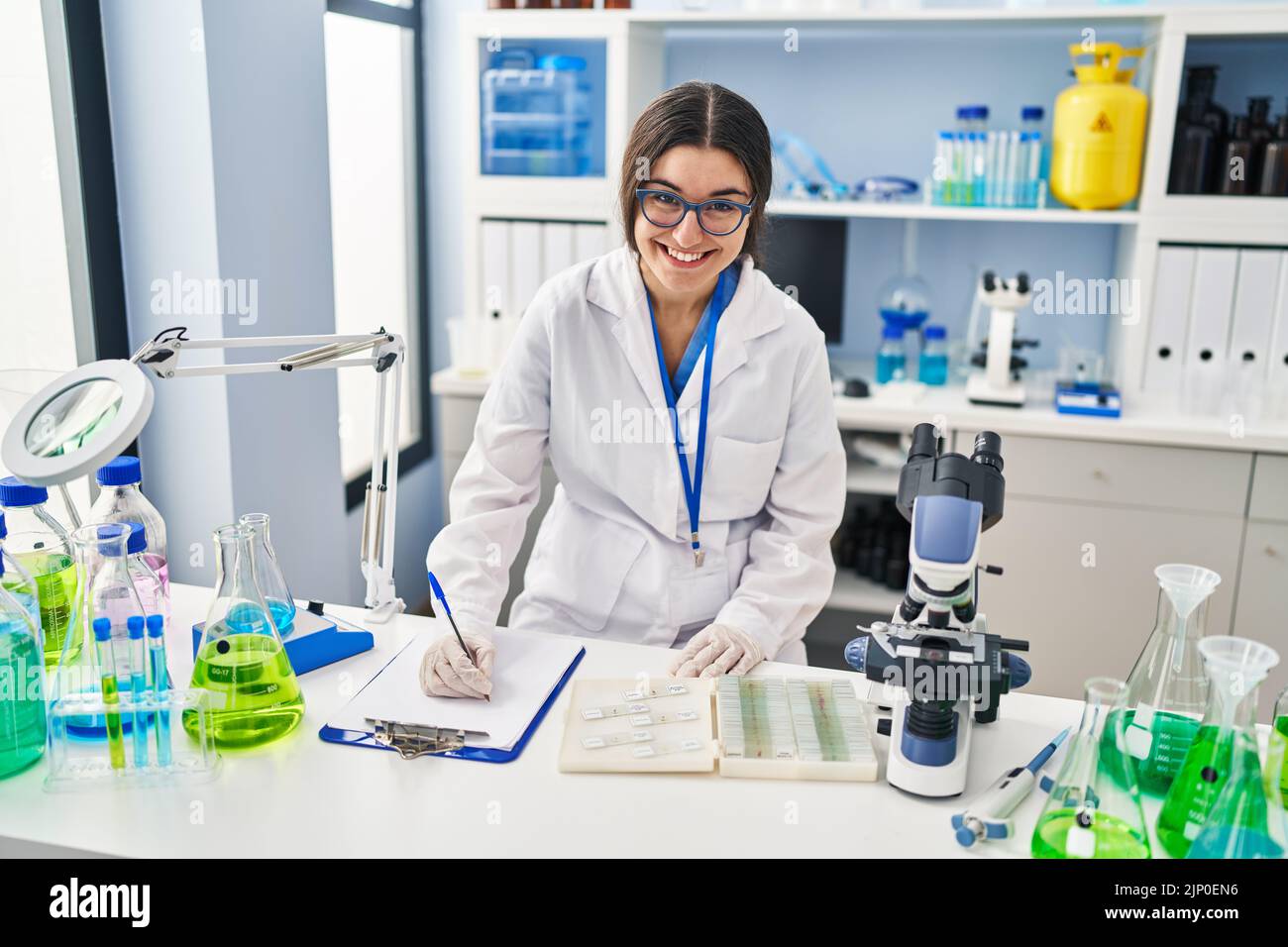 Young hispanic woman wearing scientist uniform writing on clipboard ...