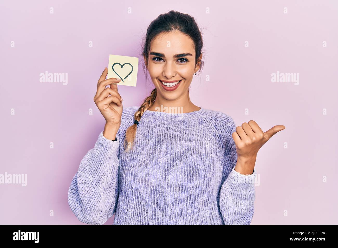 Young hispanic girl holding heart reminder pointing thumb up to the ...