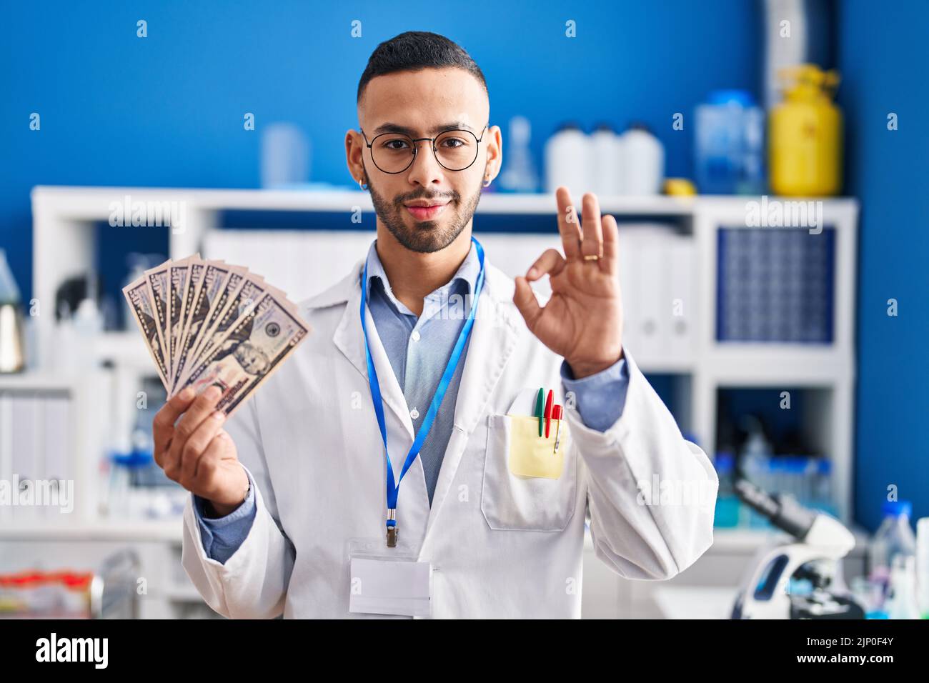 Young hispanic man working at scientist laboratory holding money doing ok sign with fingers ...