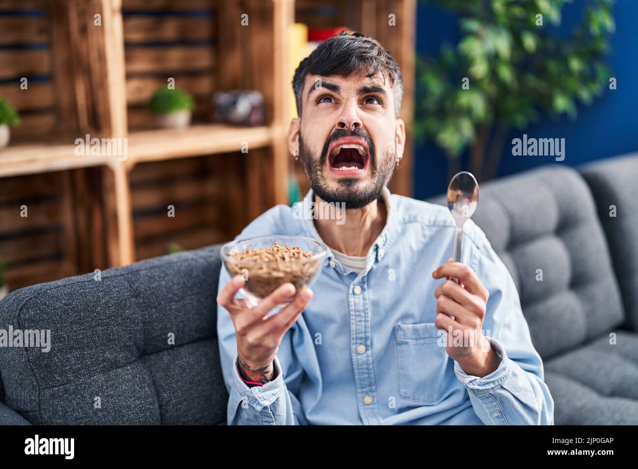 Young hispanic man with beard eating healthy whole grain cereals angry ...