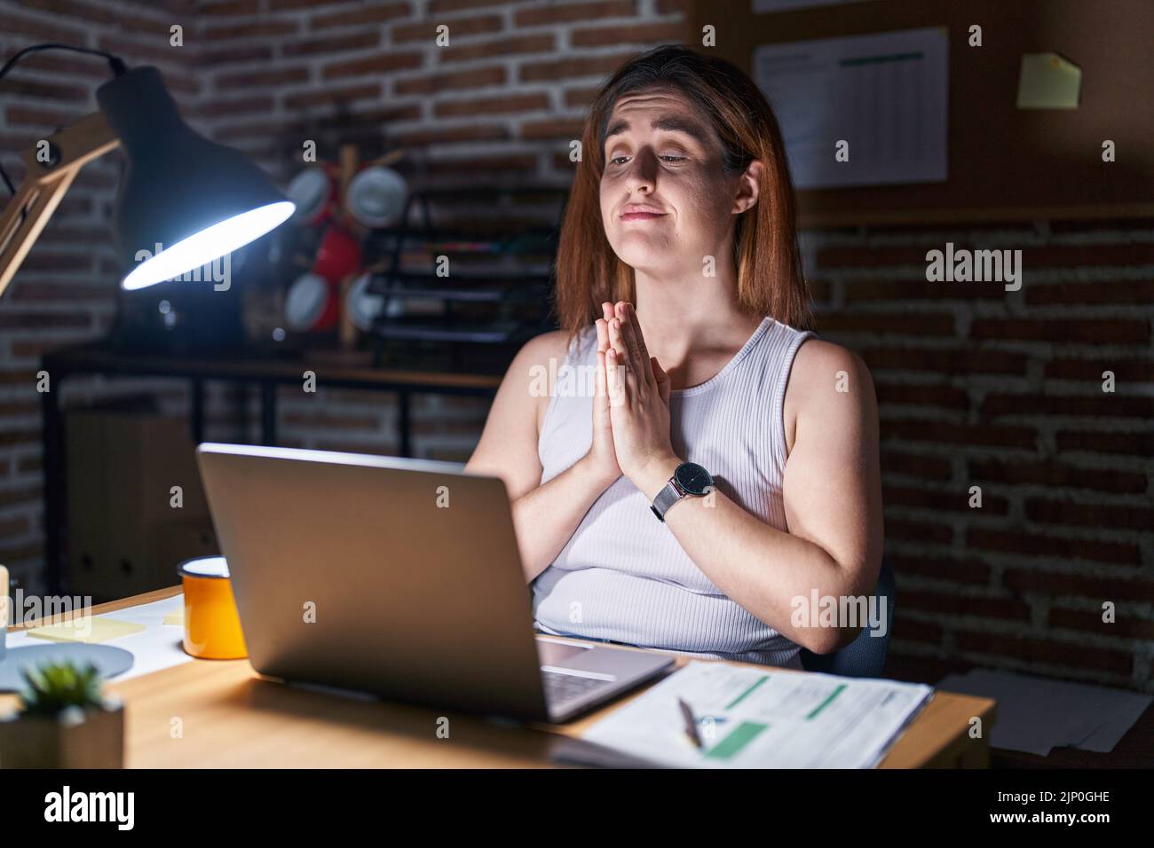Brunette woman working at the office at night praying with hands ...