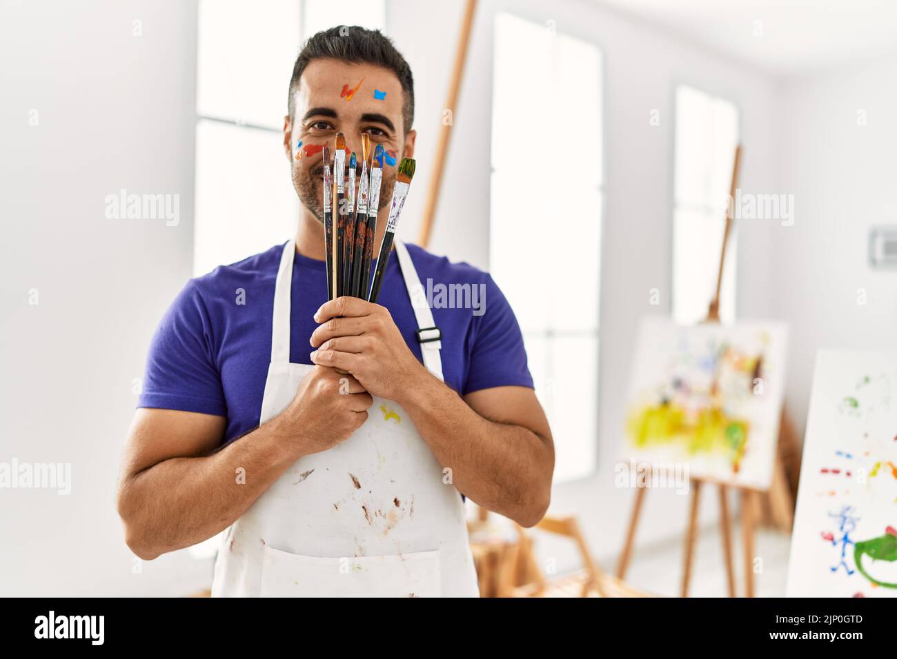 Young hispanic man smiling confident covering mouth with paintbrushes ...