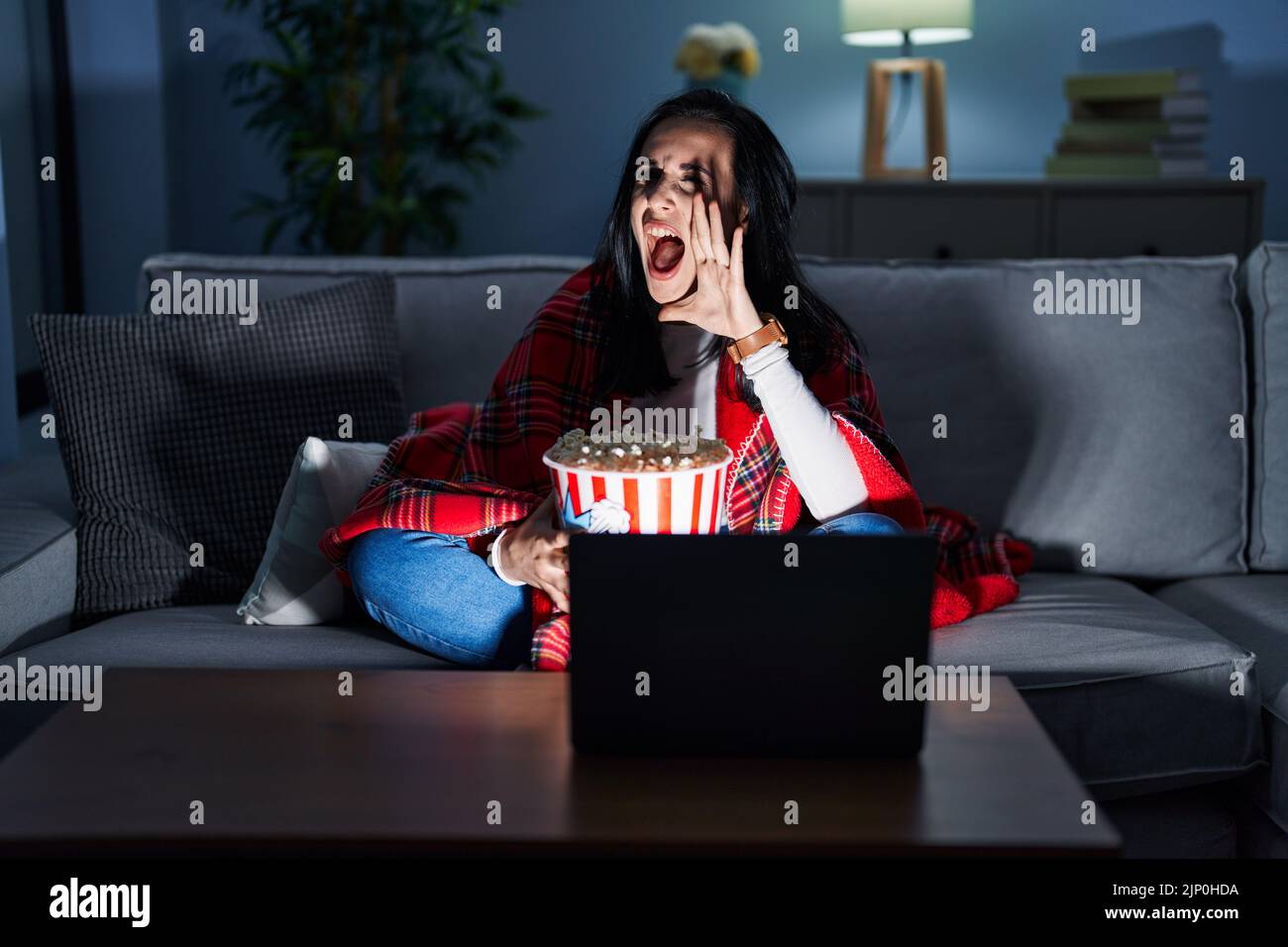 Hispanic woman eating popcorn watching a movie on the sofa shouting and ...
