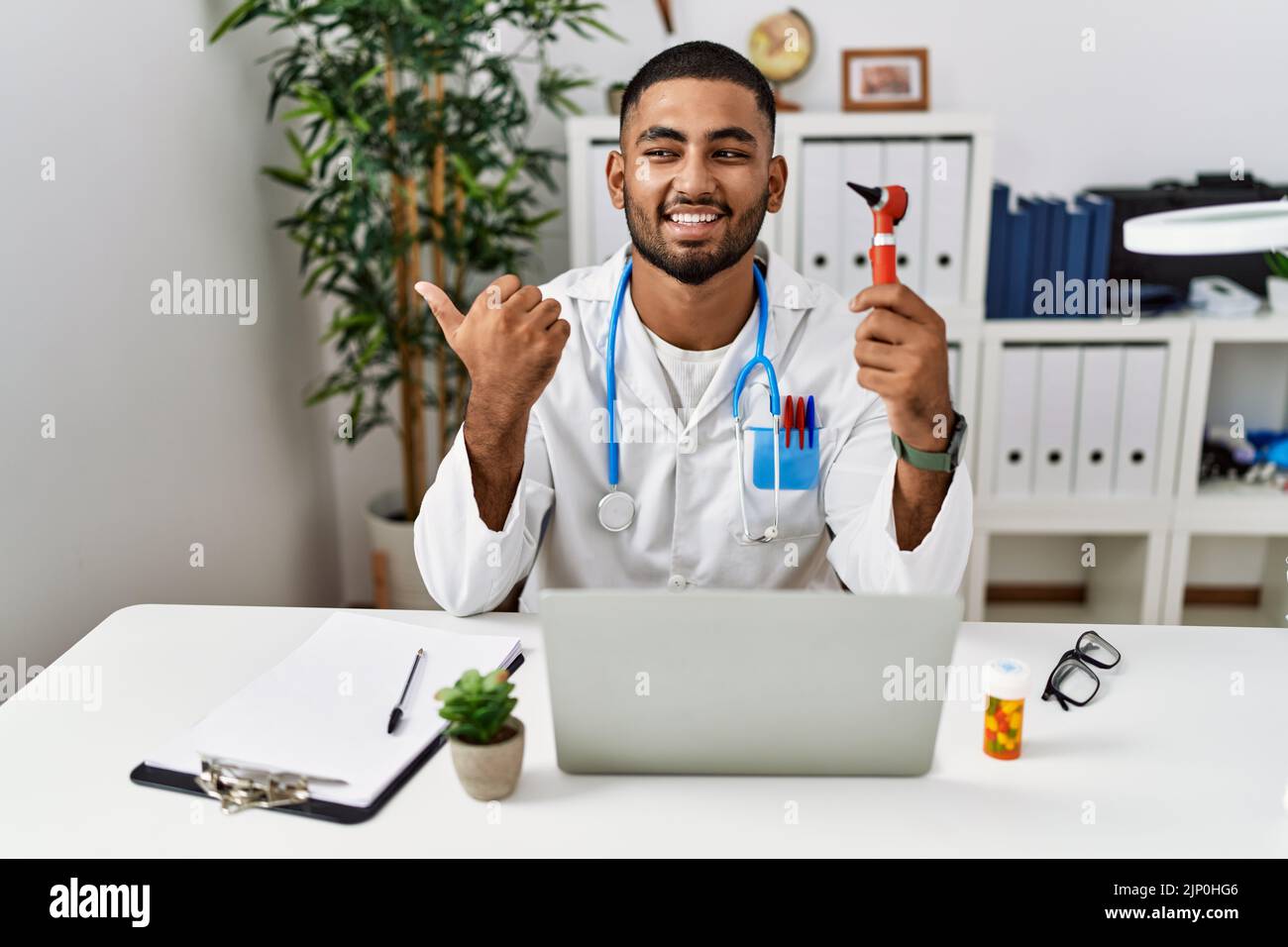 Young indian doctor holding ear otoscope pointing thumb up to the side ...