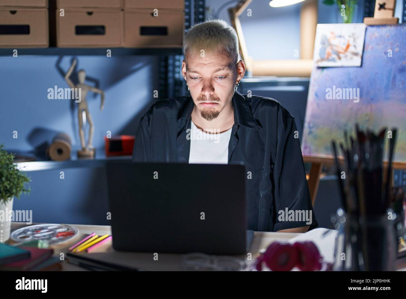 Young caucasian man using laptop at night at art studio depressed and ...