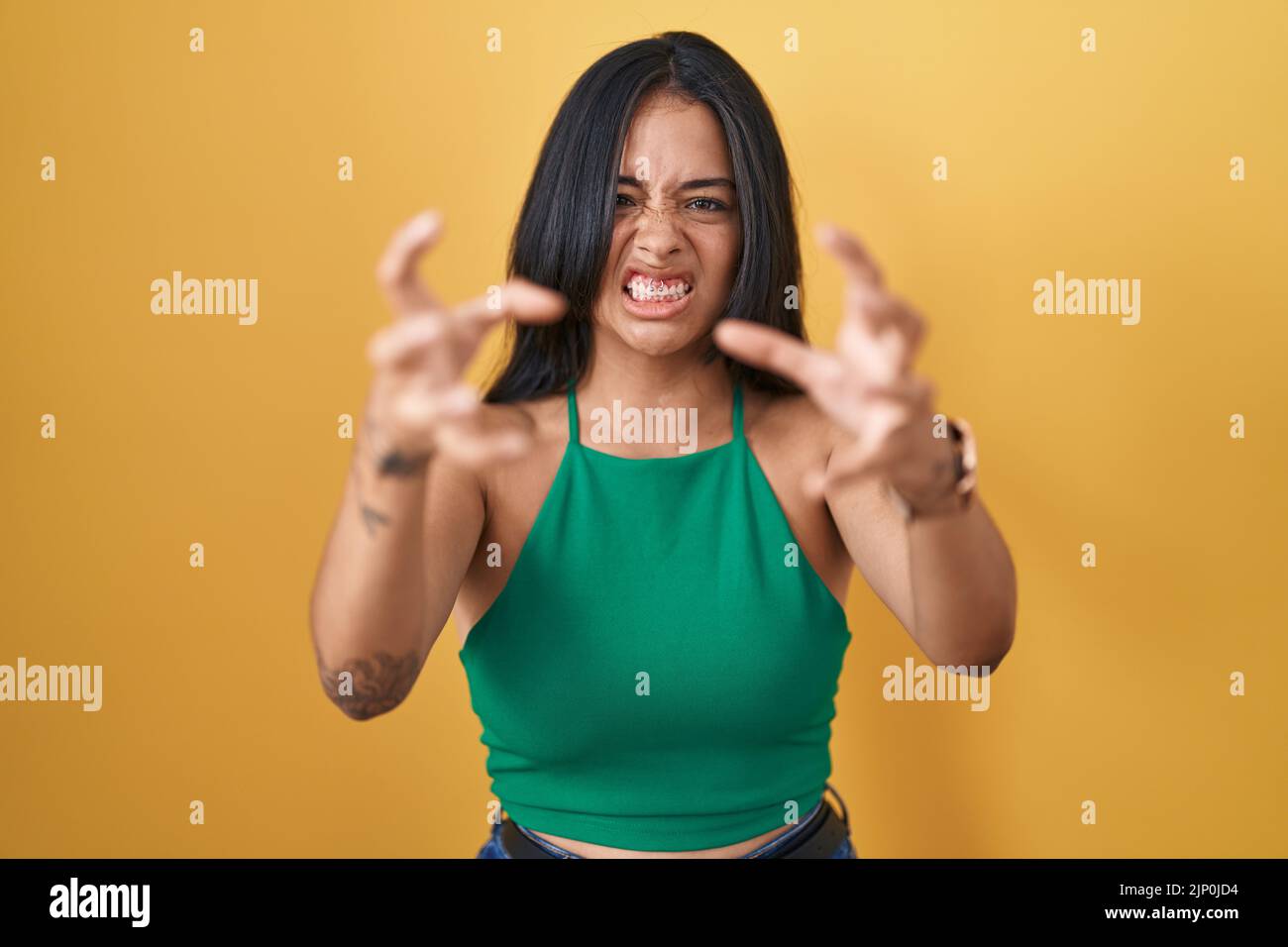 Brunette woman standing over yellow background shouting frustrated with ...