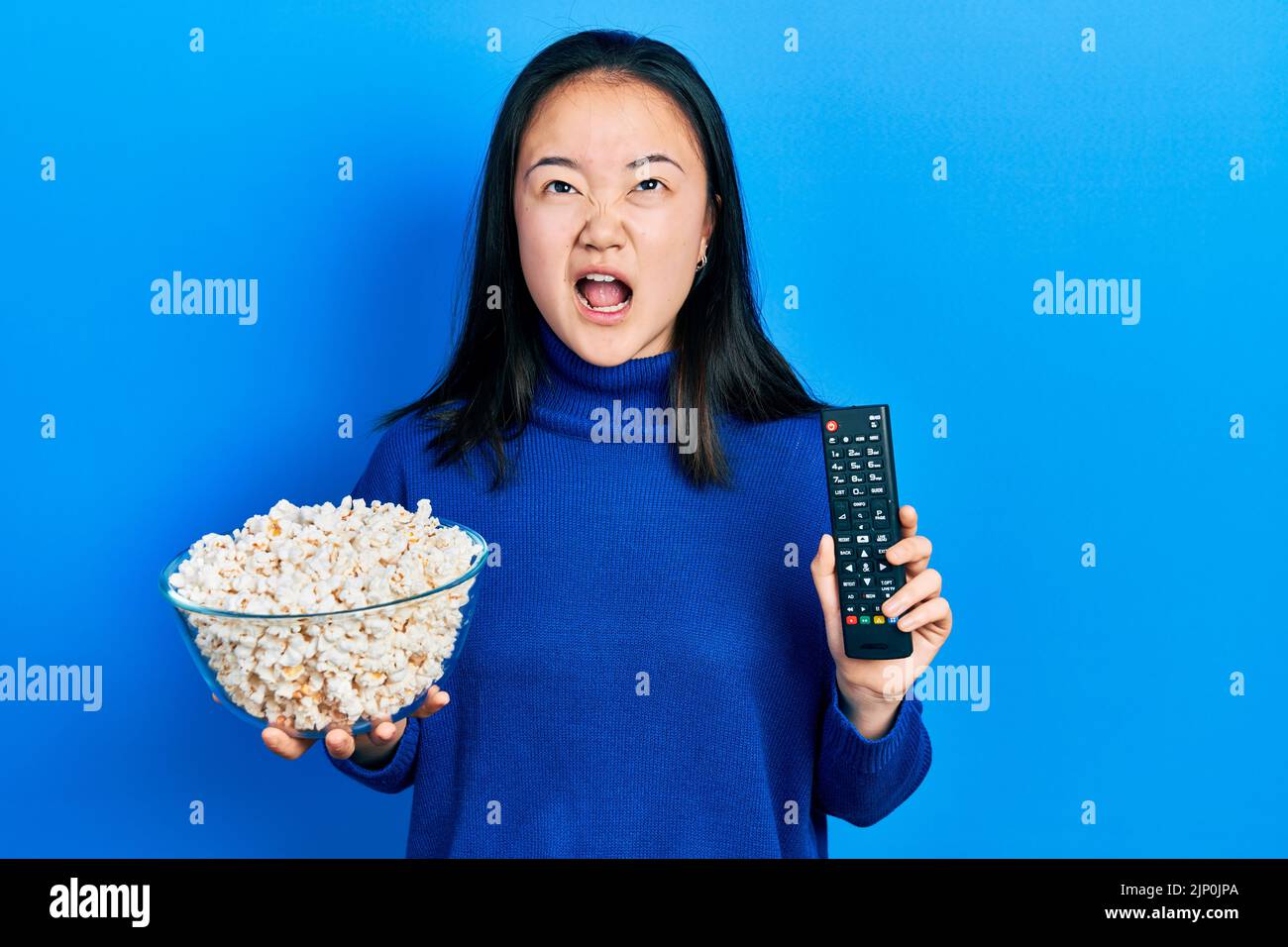 Young chinese girl eating popcorn using tv control angry and mad ...