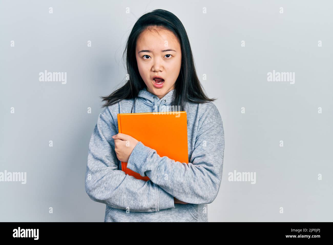 Young chinese girl holding book in shock face, looking skeptical and ...