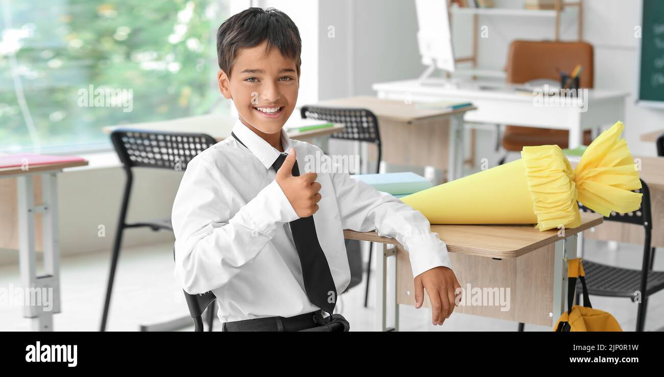 Happy little boy with school cone showing thumb-up gesture in classroom ...